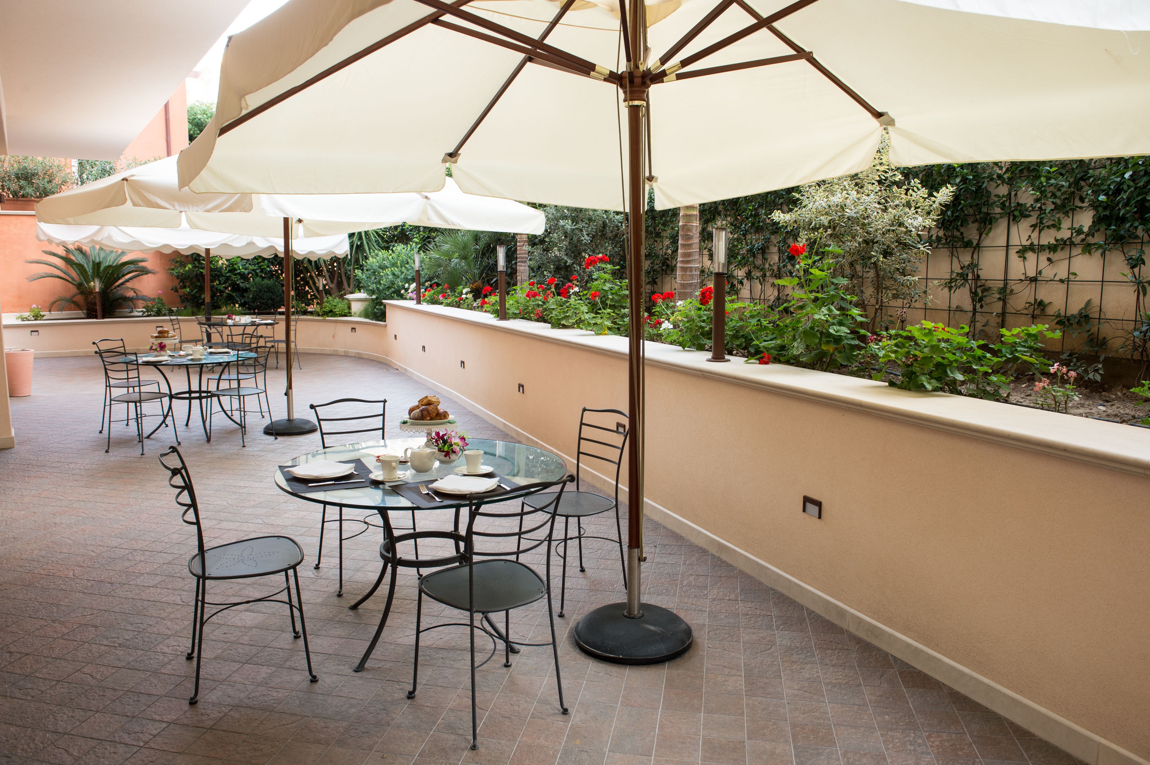 Um terraço ao ar livre com mesas e cadeiras redondas, coberto por grandes guarda-sóis, rodeado por vegetação exuberante e plantas floridas.