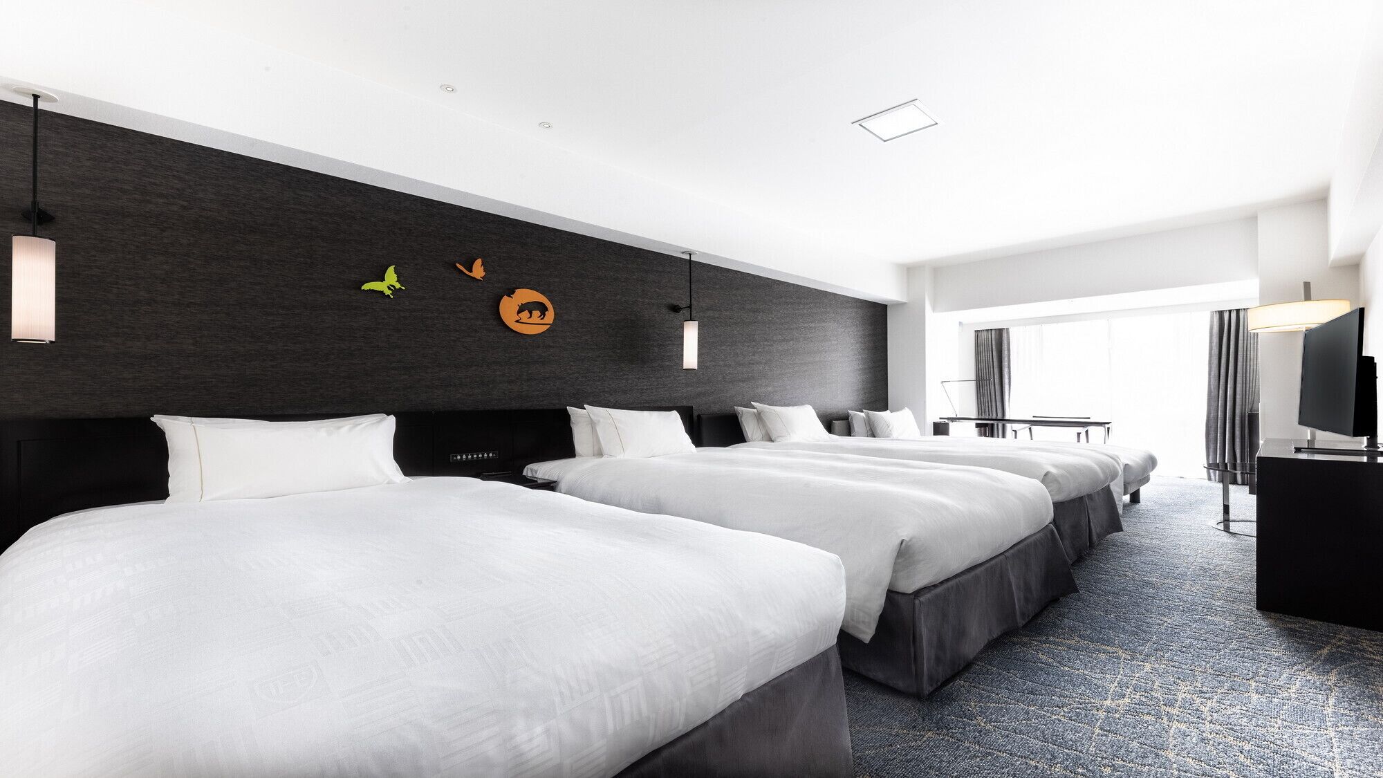 Quarto de hotel com três camas, uma escrivaninha, uma televisão e uma janela grande.