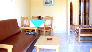 Uma zona de refeições com uma mesa posta para quatro pessoas, um sofá castanho e uma pequena mesa com uma tigela.