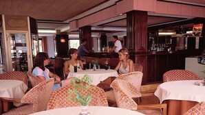 Um restaurante com mesas redondas e cadeiras de vime, um espaço de bar e vista para a cozinha.