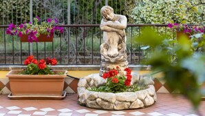 Uma estátua de pedra retrata uma figura abraçando uma criança, emoldurada por uma borda de pedra e vasos com flores vermelhas.