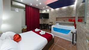 Quarto de hotel com cama, cortina vermelha, escrivaninha com cadeira, televisão, janela grande com decoração de corações vermelhos e hidromassagem.