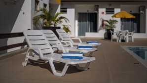 Área de piscina com espreguiçadeiras brancas e um guarda-sol amarelo.