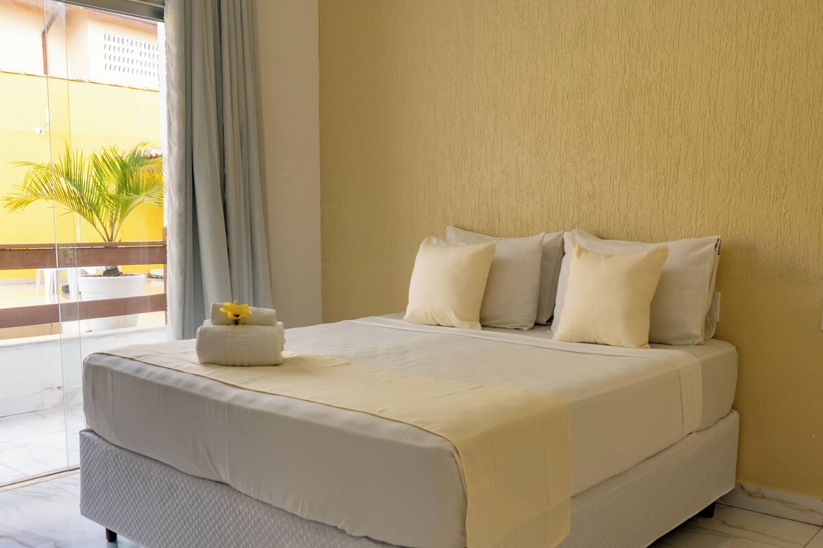 Cama bem arrumada com travesseiros e cobertor, um vaso de planta e vista para o exterior por uma janela com cortinas.