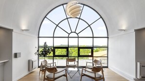 Um quarto espaçoso com uma grande janela em arco, uma mesa redonda com cadeiras e um moderno candeeiro de teto.