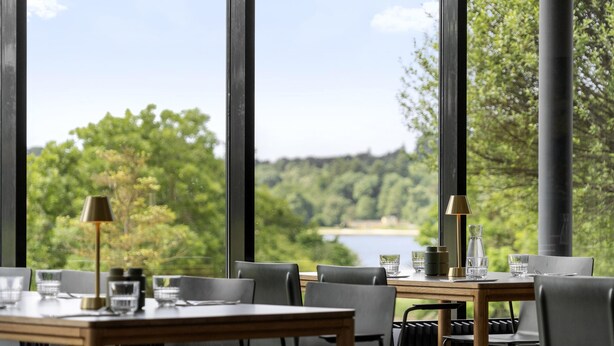 Uma área de refeições com uma mesa posta para dois, grandes janelas e vista para um lago e uma floresta.