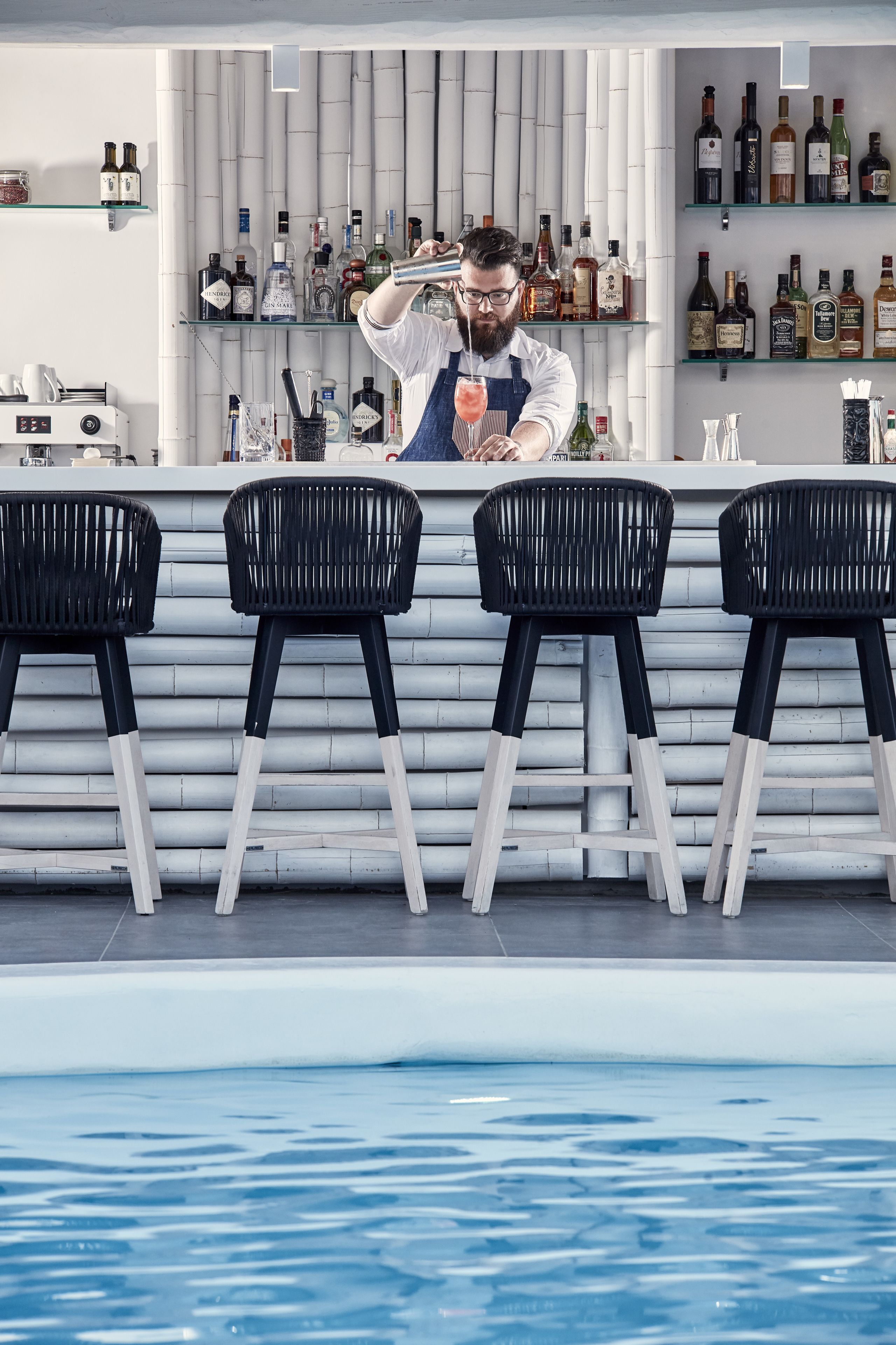 Uma área de bar com um bartender preparando uma bebida, uma piscina e banquetas de bar.