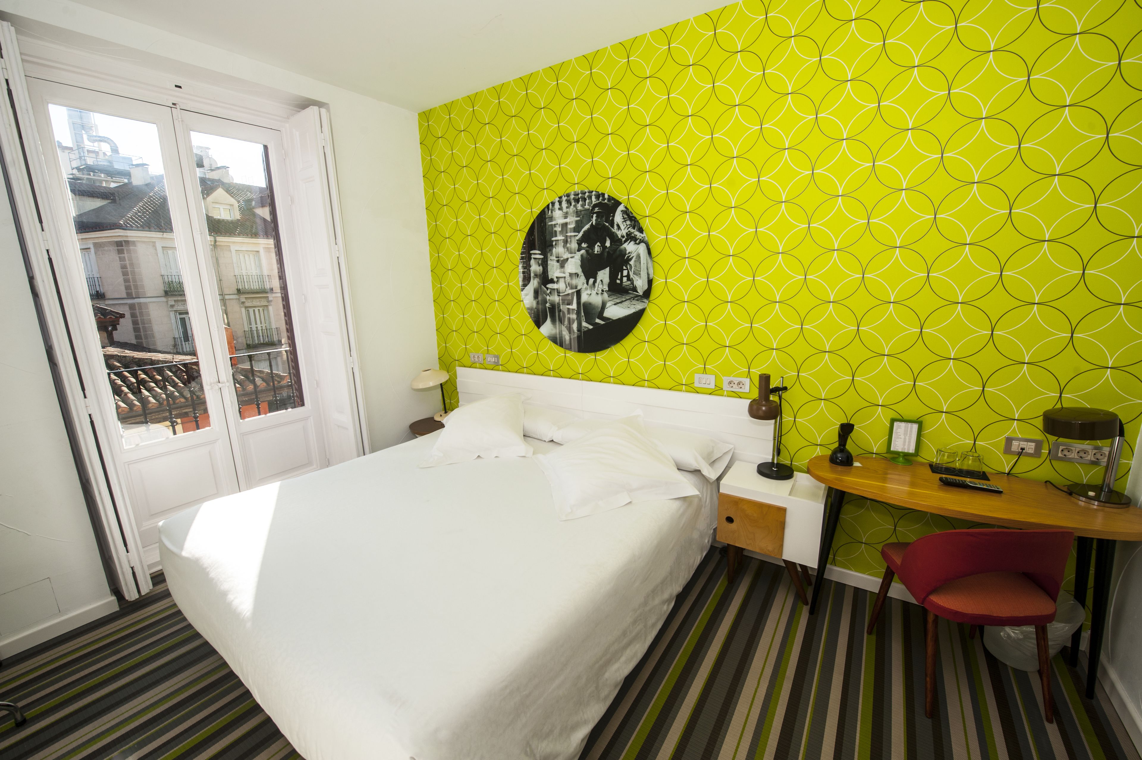 Quarto de hotel com cama, escrivaninha, cadeira, parede com padrão geométrico e janela com vista para edifícios.