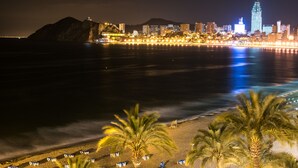 Paisagem costeira noturna com edifícios iluminados, uma praia com palmeiras e uma montanha ao fundo.