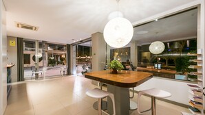 Um hall de hotel moderno com uma mesa de madeira central, dois bancos altos, uma planta em vaso e uma grande luminária pendente.
