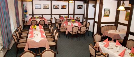 Uma sala de jantar com várias mesas preparadas para o serviço, com decoração em estilo europeu tradicional, incluindo vigas de madeira aparentes e luminárias pendentes.
