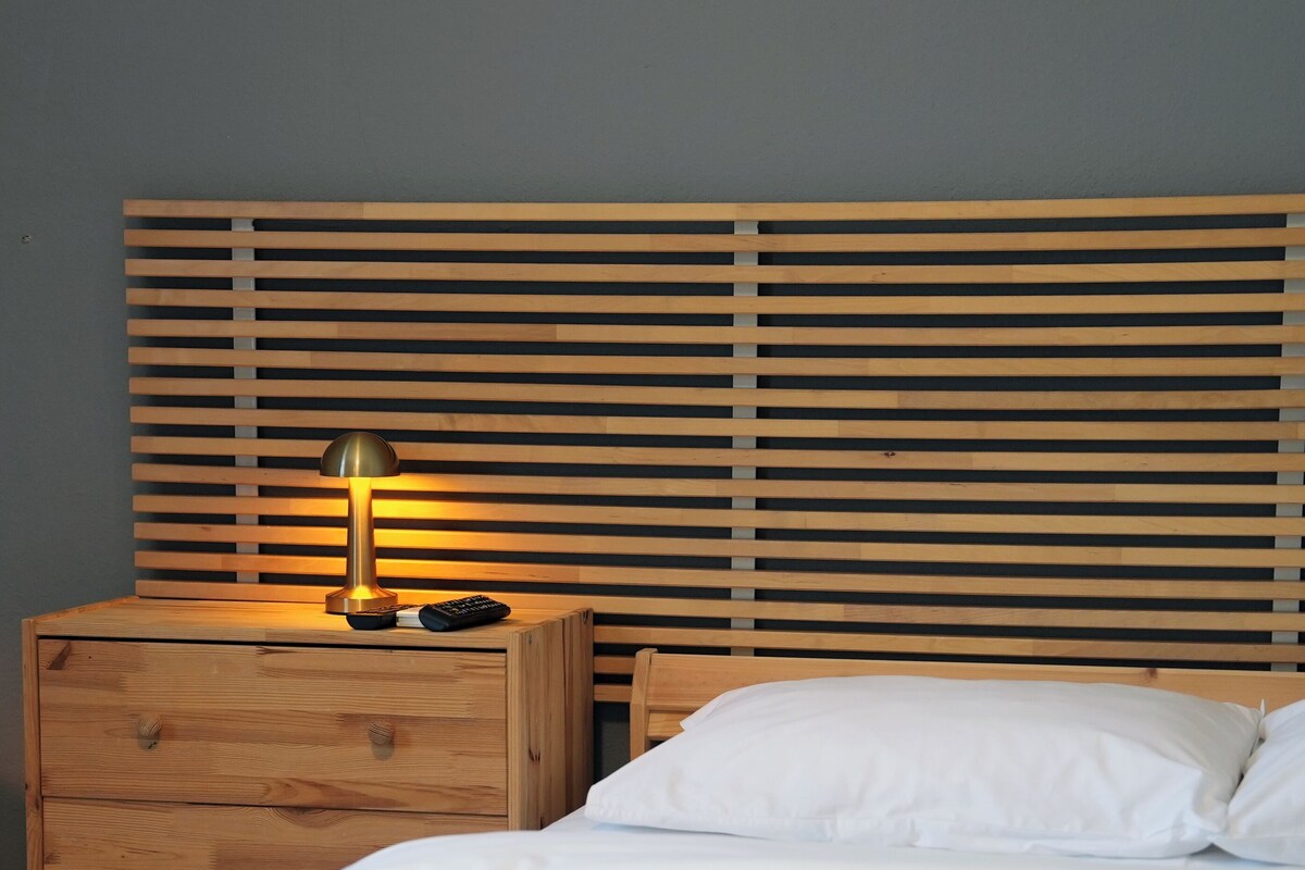 Cama bem arrumada com lençóis brancos, mesa de cabeceira de madeira com abajur, e cabeceira de madeira com ripas horizontais.