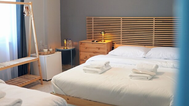 Quarto de hotel com cama, cabeceira de madeira, mesa de cabeceira com luminária, pequena mesa com garrafas e prateleira de madeira.