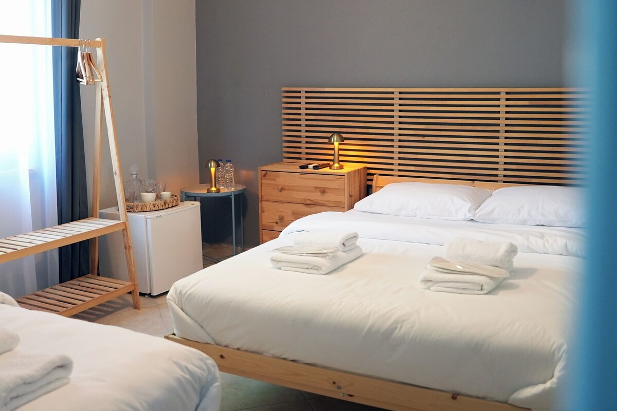 Quarto de hotel com cama, cabeceira de madeira, mesa de cabeceira com luminária, pequena mesa com garrafas e prateleira de madeira.