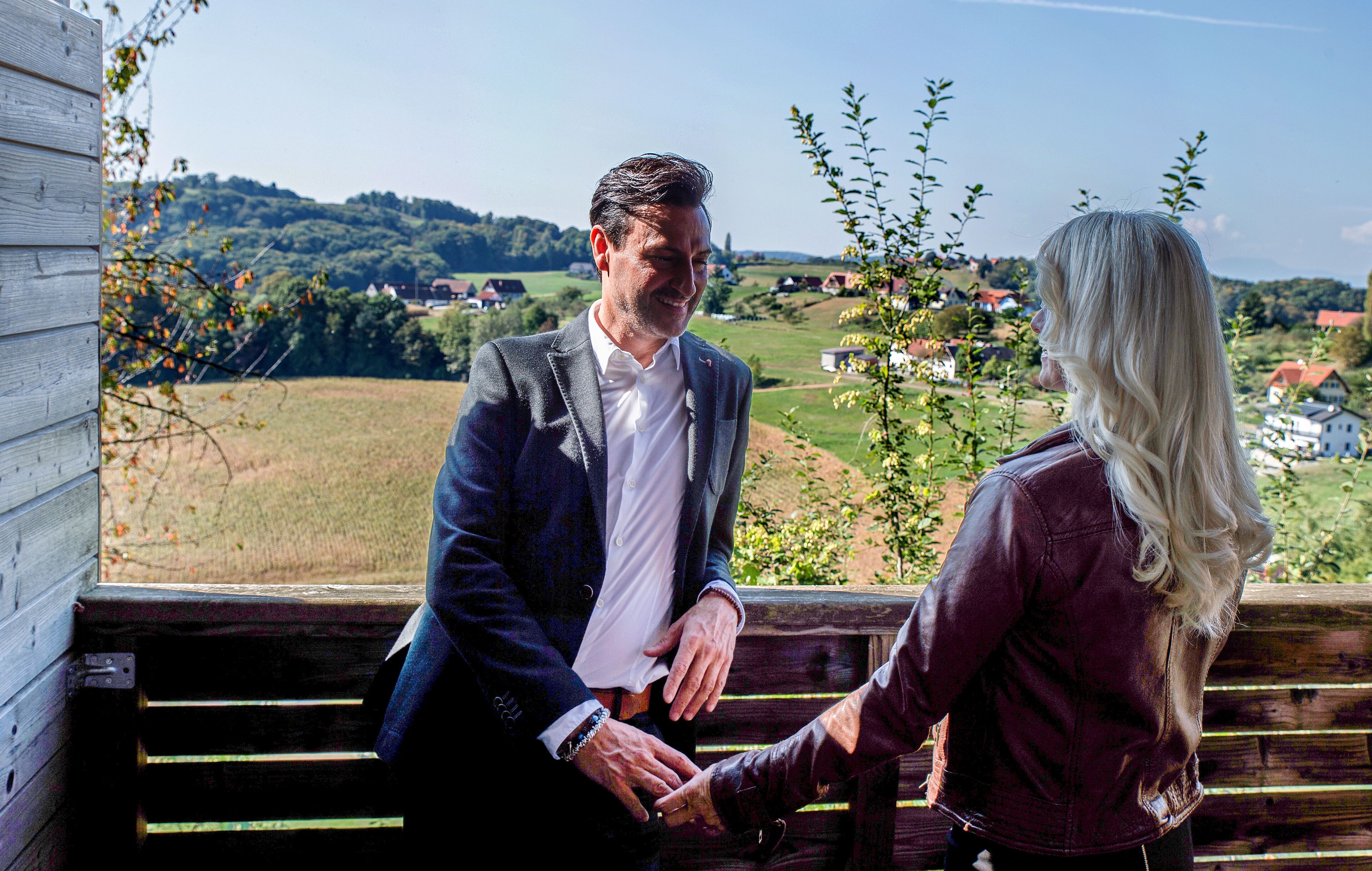 Um casal sentado em um banco de madeira com uma vista panorâmica de uma aldeia e colinas ao fundo.