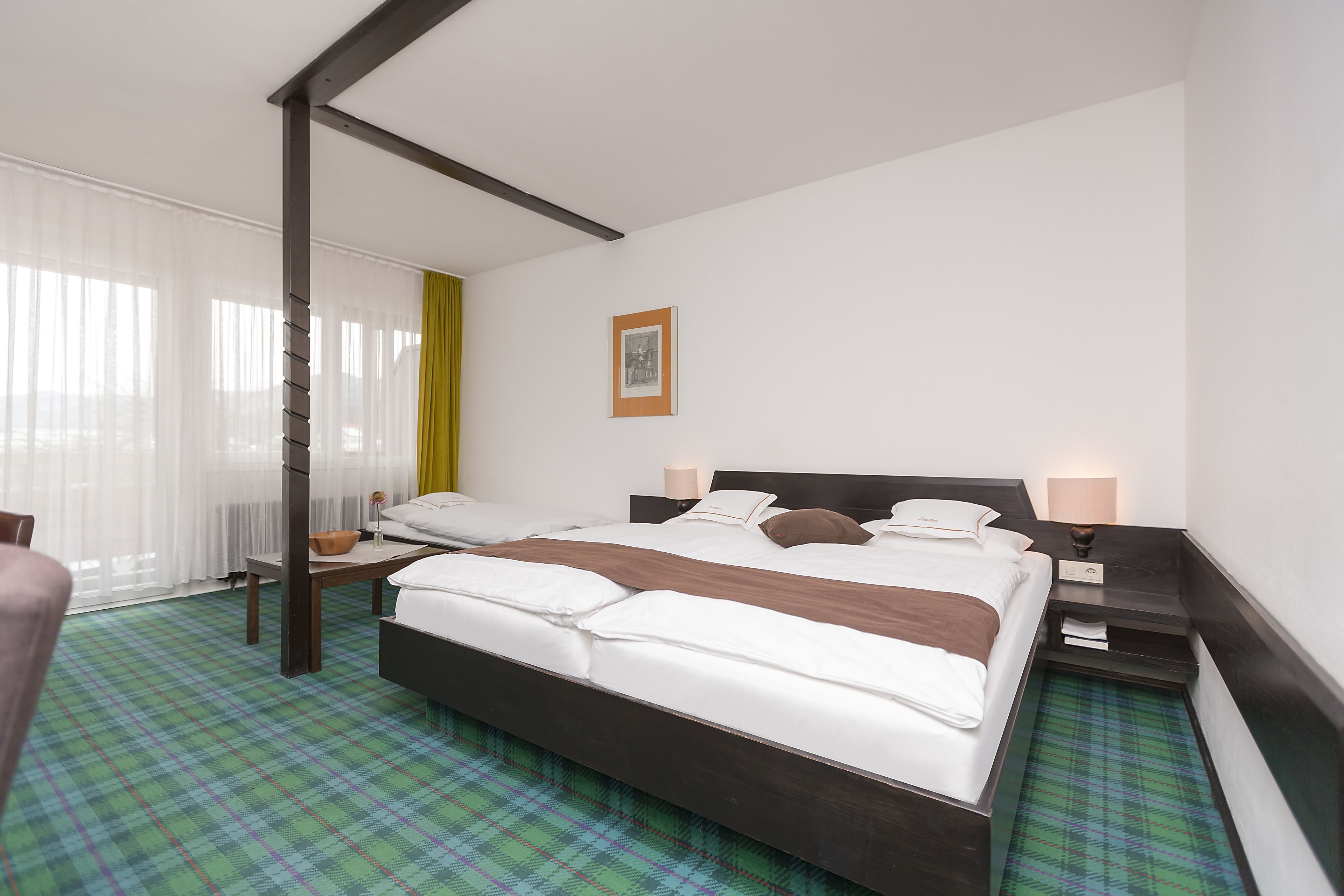 Quarto de hotel com uma cama grande, mesa de cabeceira, cadeira e janela com cortinas leves.