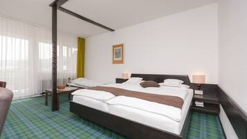 Quarto de hotel com uma cama grande, mesa de cabeceira, cadeira e janela com cortinas leves.