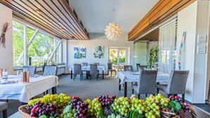 Uma área de refeições com mesas preparadas para uma refeição, uma grande tigela com uvas variadas em primeiro plano e vista para o exterior através de grandes janelas.