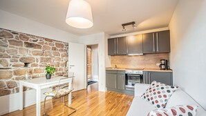 Uma cozinha moderna com armários cinza e bancada de madeira, uma sala de jantar com mesa branca e cadeiras de metal, e uma sala de estar com sofá branco e almofadas estampadas.