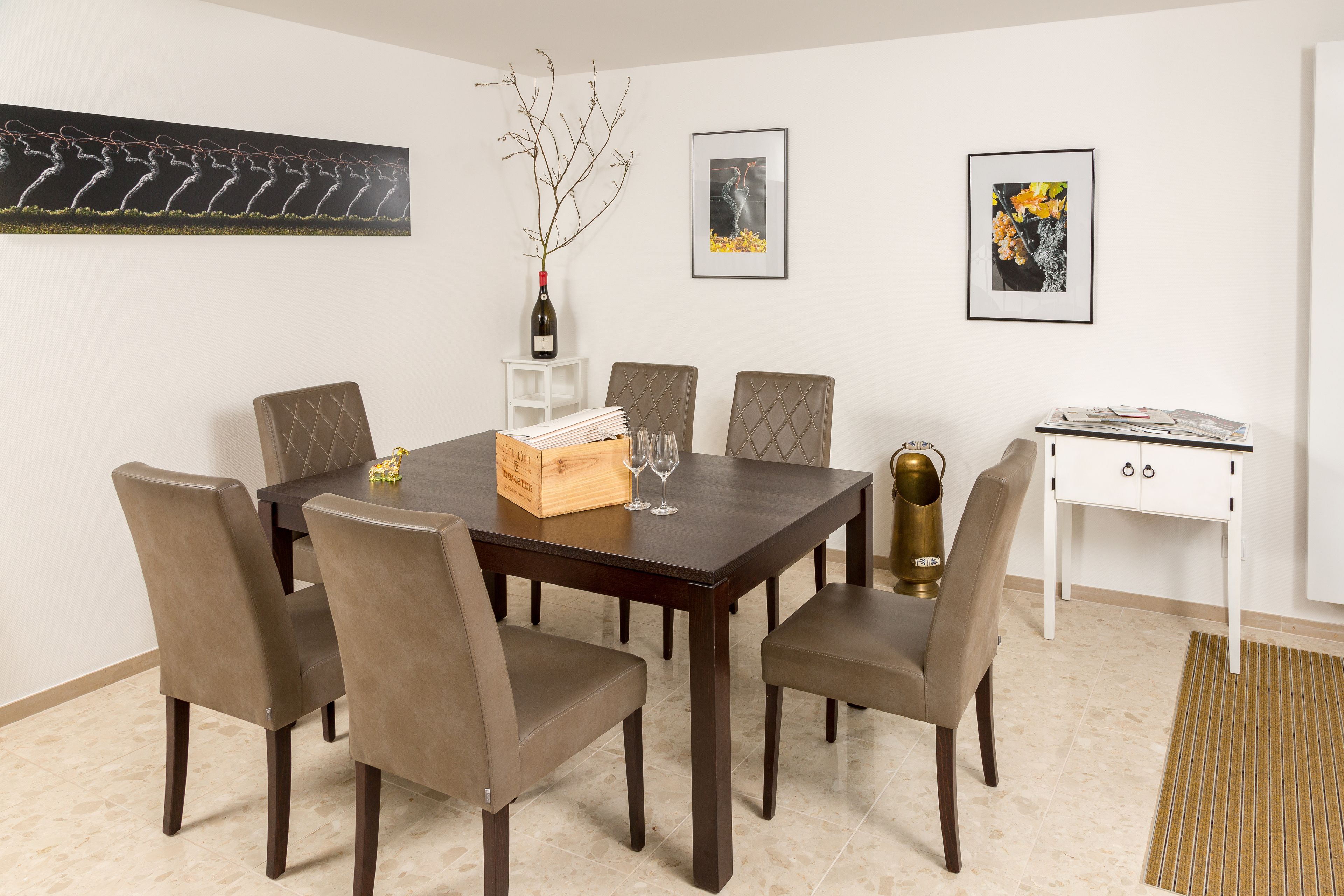 Uma sala de jantar com uma mesa de madeira escura, seis cadeiras, um vaso com galhos e quadros nas paredes.