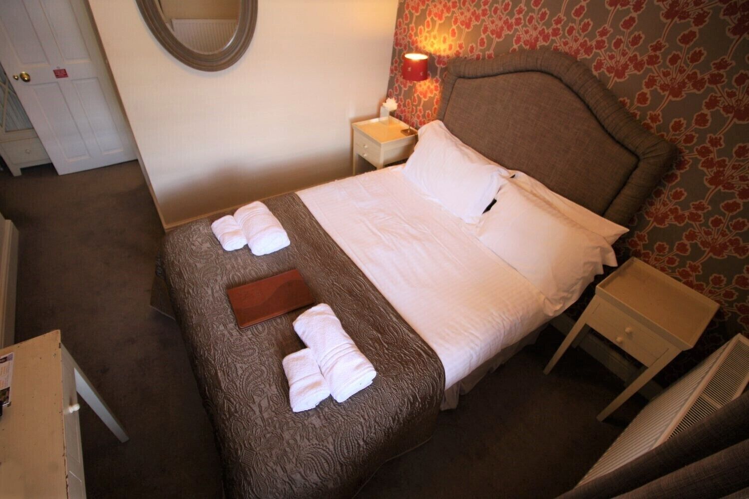 Cama bem arrumada com lençóis brancos e duas toalhas dobradas. Apresenta uma mesa de cabeceira com abajur e um pequeno tabuleiro. O quarto possui papel de parede floral e uma porta ao fundo.