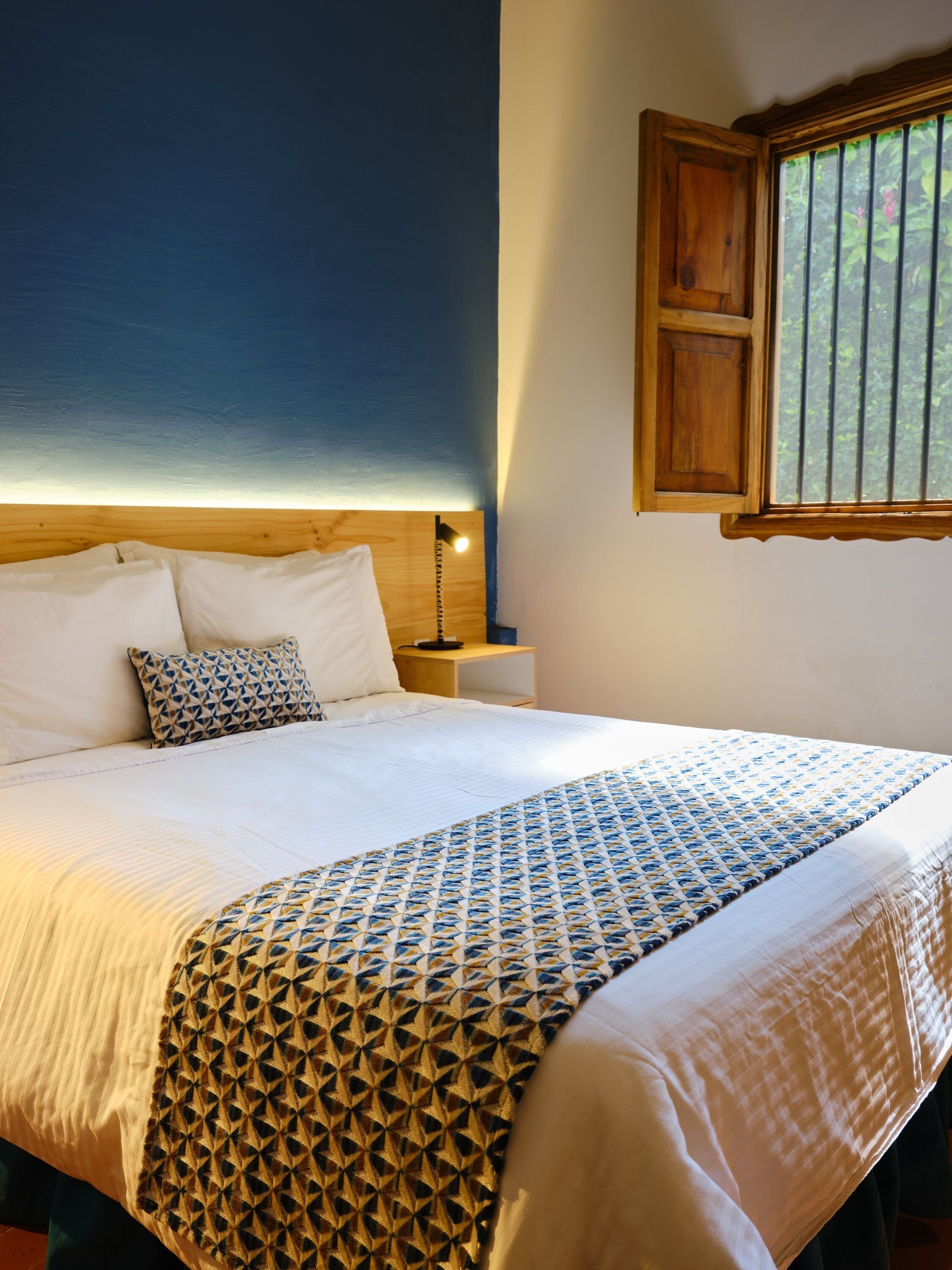 Uma cama bem arrumada com colcha estampada, abajur de cabeceira e janela com moldura de madeira.