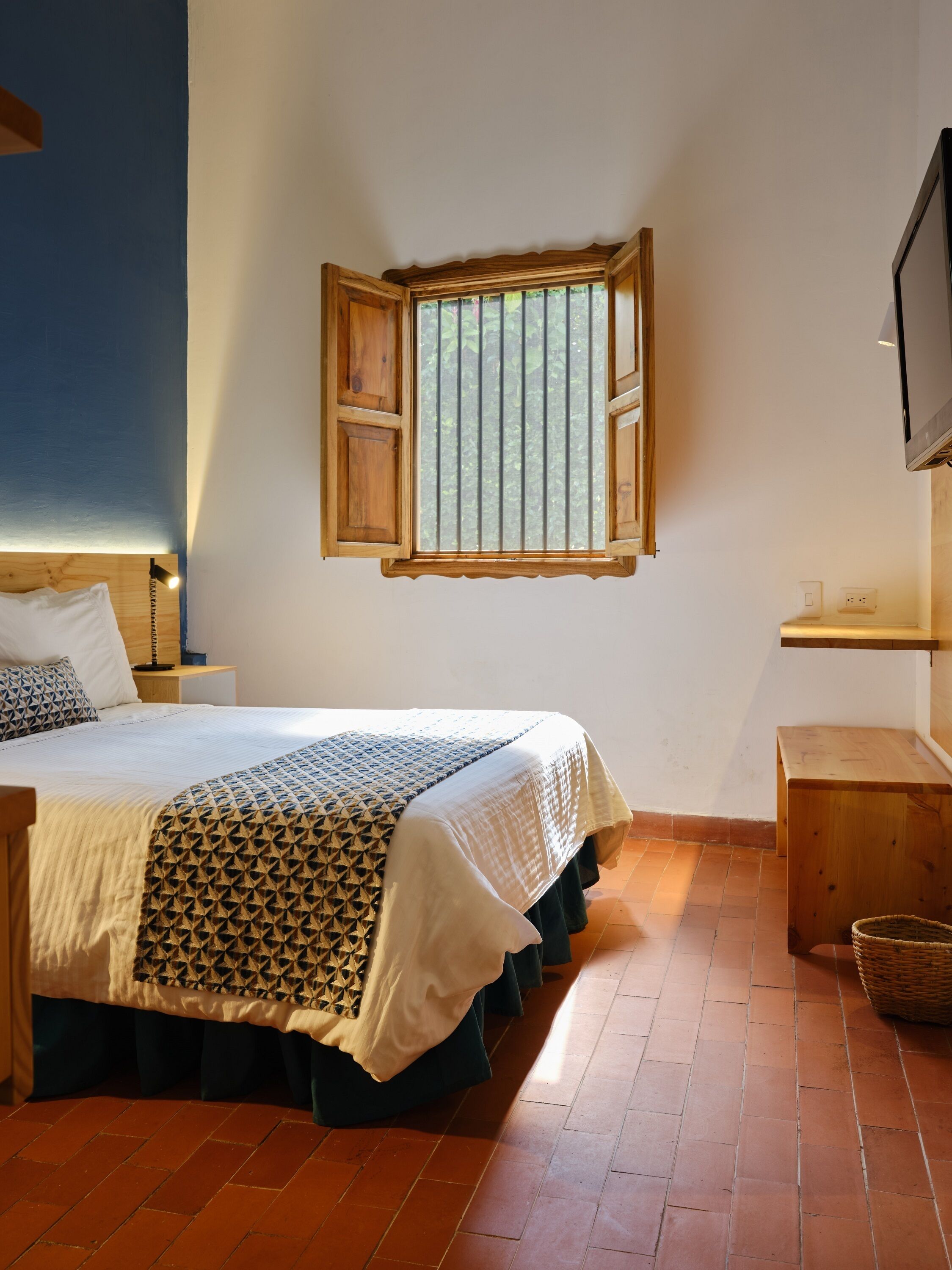 Uma cama bem arrumada com colcha estampada, uma mesinha de cabeceira de madeira com abajur, uma pequena mesa de madeira e uma janela com persianas de madeira.