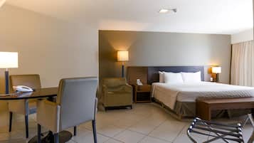Quarto de hotel com cama, duas mesas de cabeceira com abajures, uma escrivaninha com cadeira e uma poltrona pequena.