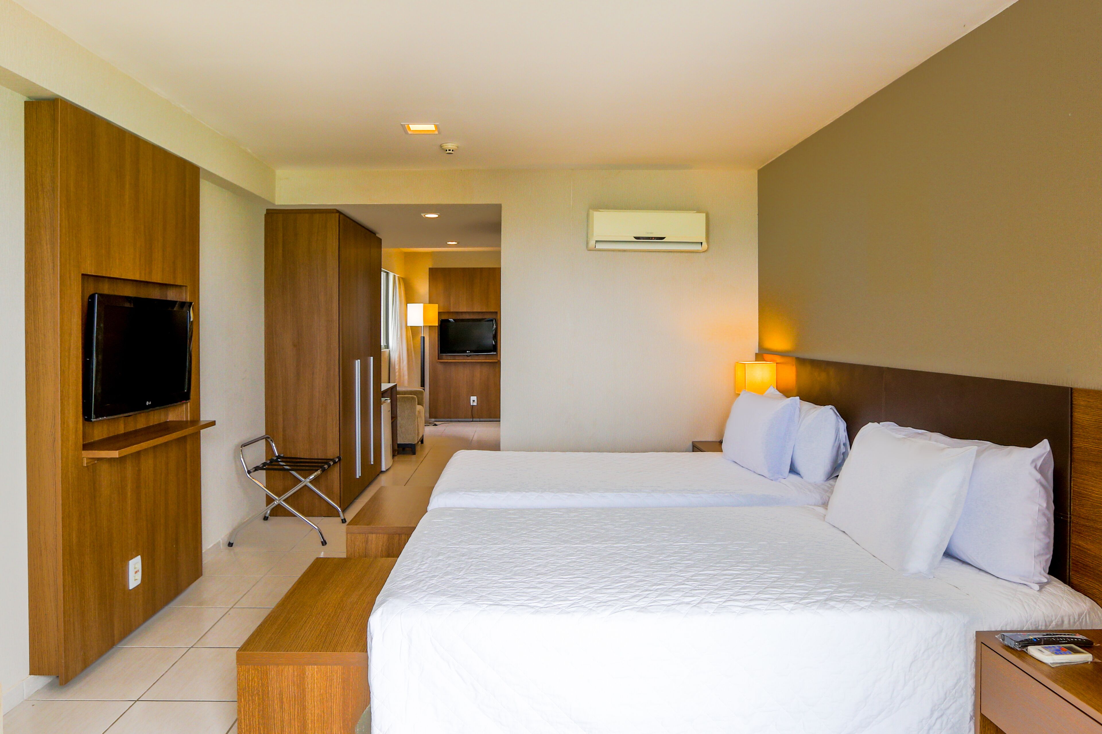 Quarto de hotel com duas camas, televisão montada na parede, televisão de tela plana, armário de madeira, mesa de trabalho com luminária e mesa de cabeceira com controle remoto.