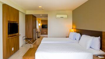 Quarto de hotel com duas camas, televisão montada na parede, televisão de tela plana, armário de madeira, mesa de trabalho com luminária e mesa de cabeceira com controle remoto.