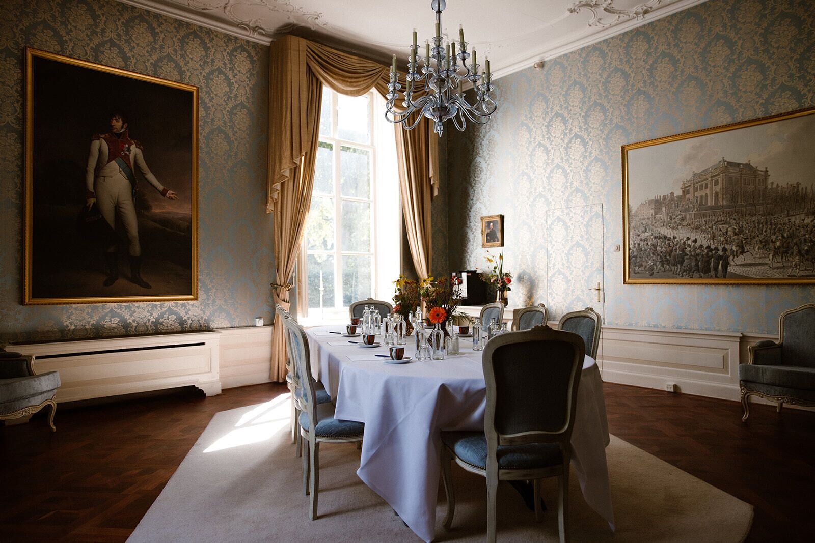 Uma sala de jantar formal com uma mesa posta para quatro pessoas, rodeada por cadeiras ricamente decoradas, um candelabro e duas grandes pinturas emolduradas.