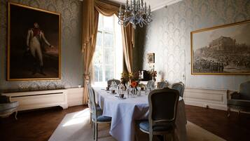 Uma sala de jantar formal com uma mesa posta para quatro pessoas, rodeada por cadeiras ricamente decoradas, um candelabro e duas grandes pinturas emolduradas.