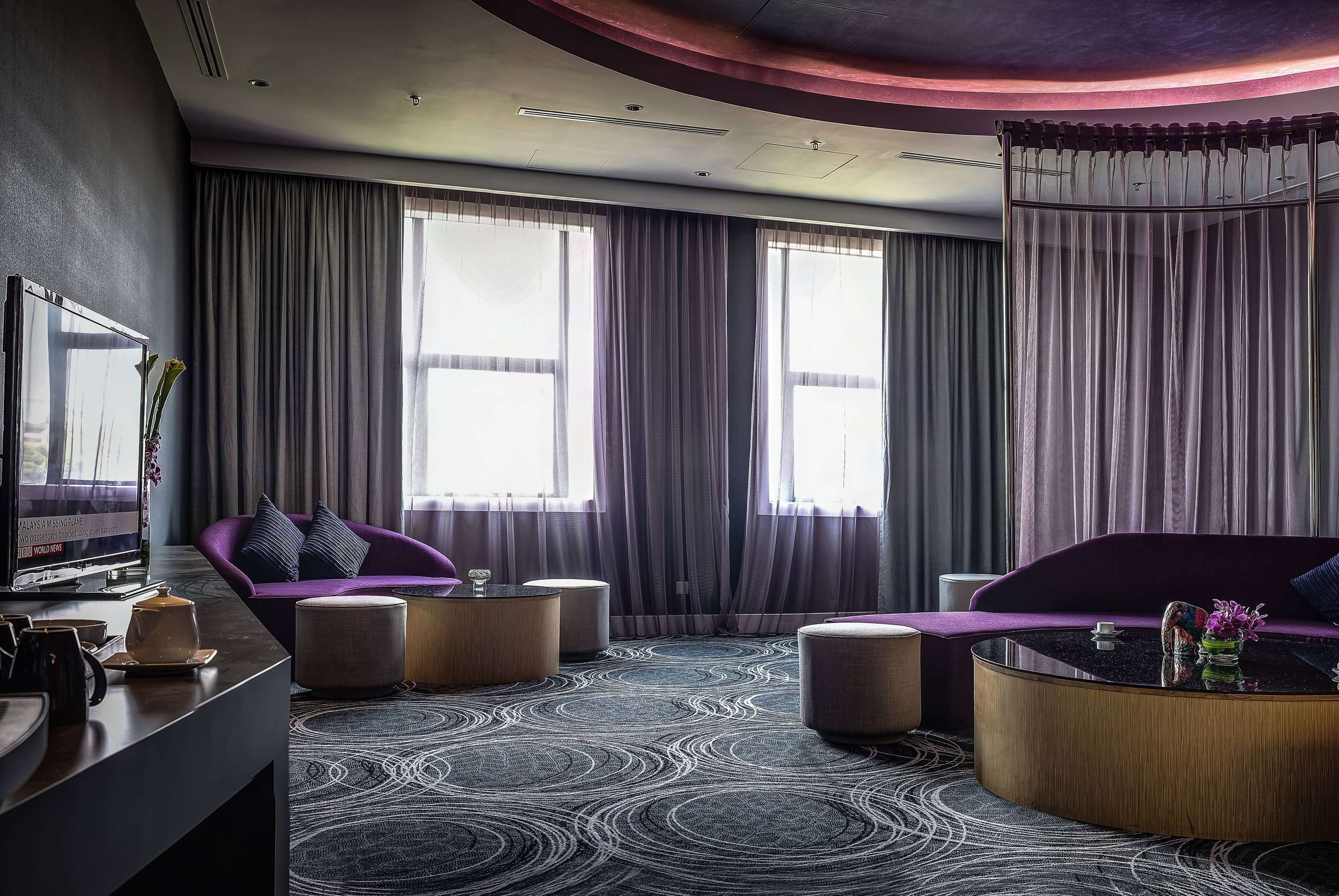 Quarto de hotel moderno com uma janela grande, televisão de tela plana, sofá, mesa de centro e carpete estampado.