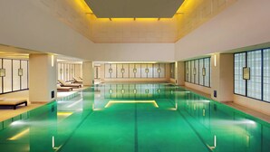 Uma grande piscina interior com piso verde, rodeada por áreas de estar e janelas de estilo tradicional.