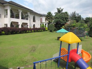 Um parquinho com escorrega e estruturas de escalada, um guarda-sol verde e um edifício com janelas em arco e sacadas ao fundo.