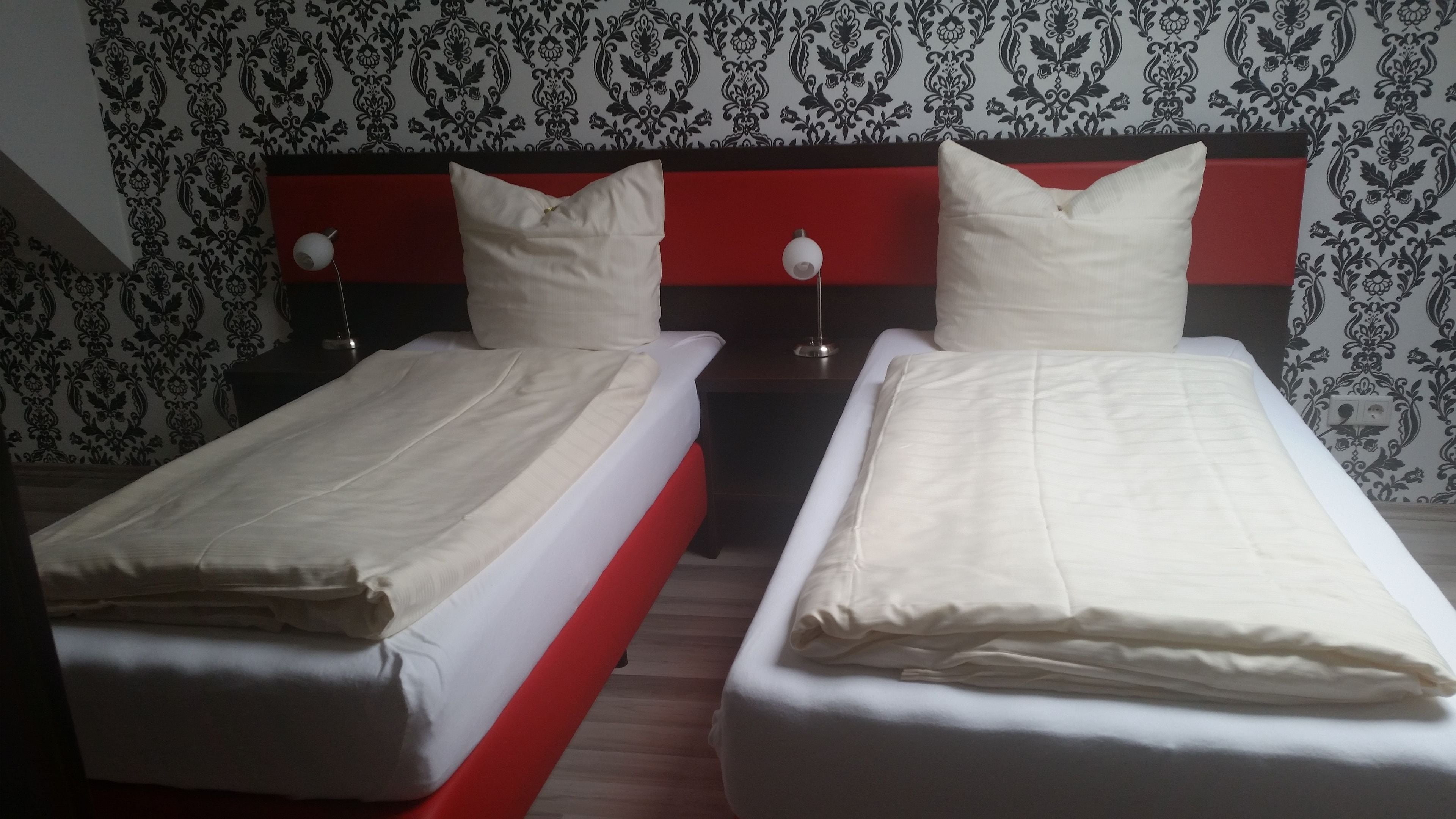 Duas camas de solteiro com cabeceiras vermelhas e roupa de cama branca, um criado-mudo com abajur e um papel de parede estampado.
