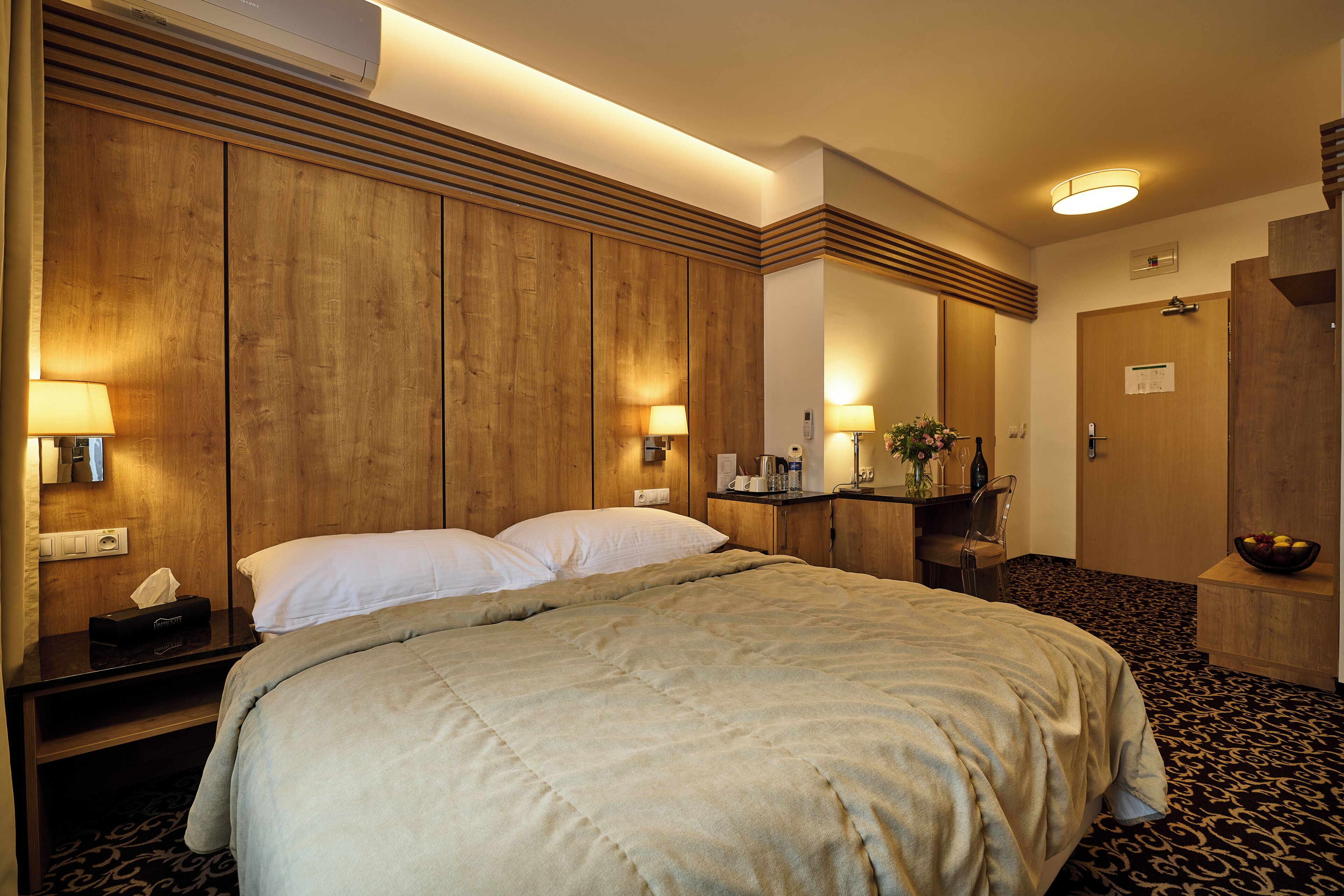 Quarto de hotel com uma cama grande, painéis de madeira, uma escrivaninha com abajur, uma mesinha com um vaso e uma porta com placa.