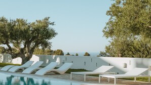 Uma área à beira da piscina com espreguiçadeiras brancas e vista para o mar.