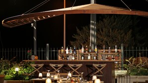 Uma área de bar ao ar livre com mesas e cadeiras de madeira, um toldo de madeira e diversos frascos e copos.