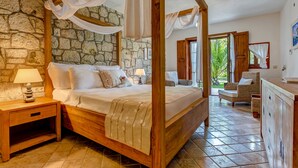 Uma cama com dossel sobre quatro colunas, com um painel de parede de pedra, uma mesa de cabeceira de madeira com abajur e uma cadeira de vime em um quarto com piso de azulejos.
