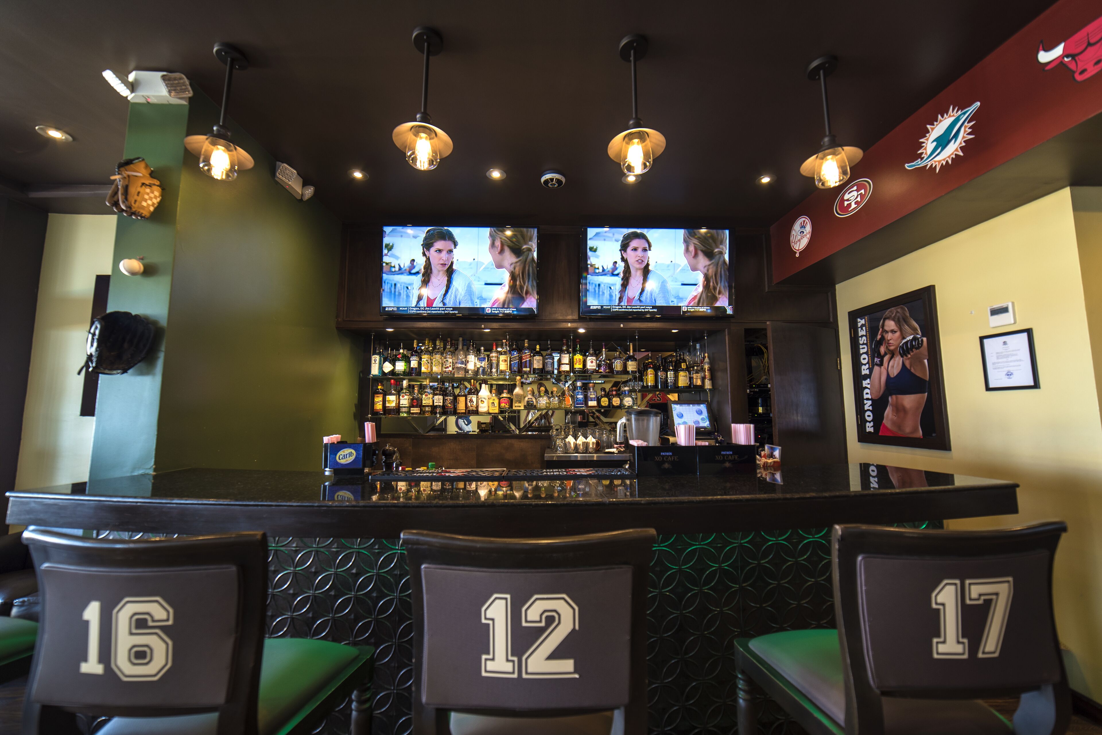 Uma área de bar com várias televisões, um bar bem abastecido e banquetas numeradas.