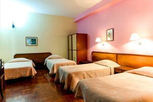 Quarto de hotel com quatro camas de solteiro, cabeceiras de madeira, guarda-roupa de madeira, mesa de cabeceira e duas luminárias fixadas na parede.