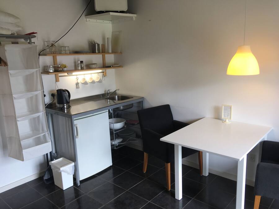 Uma cozinha compacta com pia, chaleira e um refrigerador pequeno. Há uma mesa branca com duas cadeiras e um lustre pendurado acima.