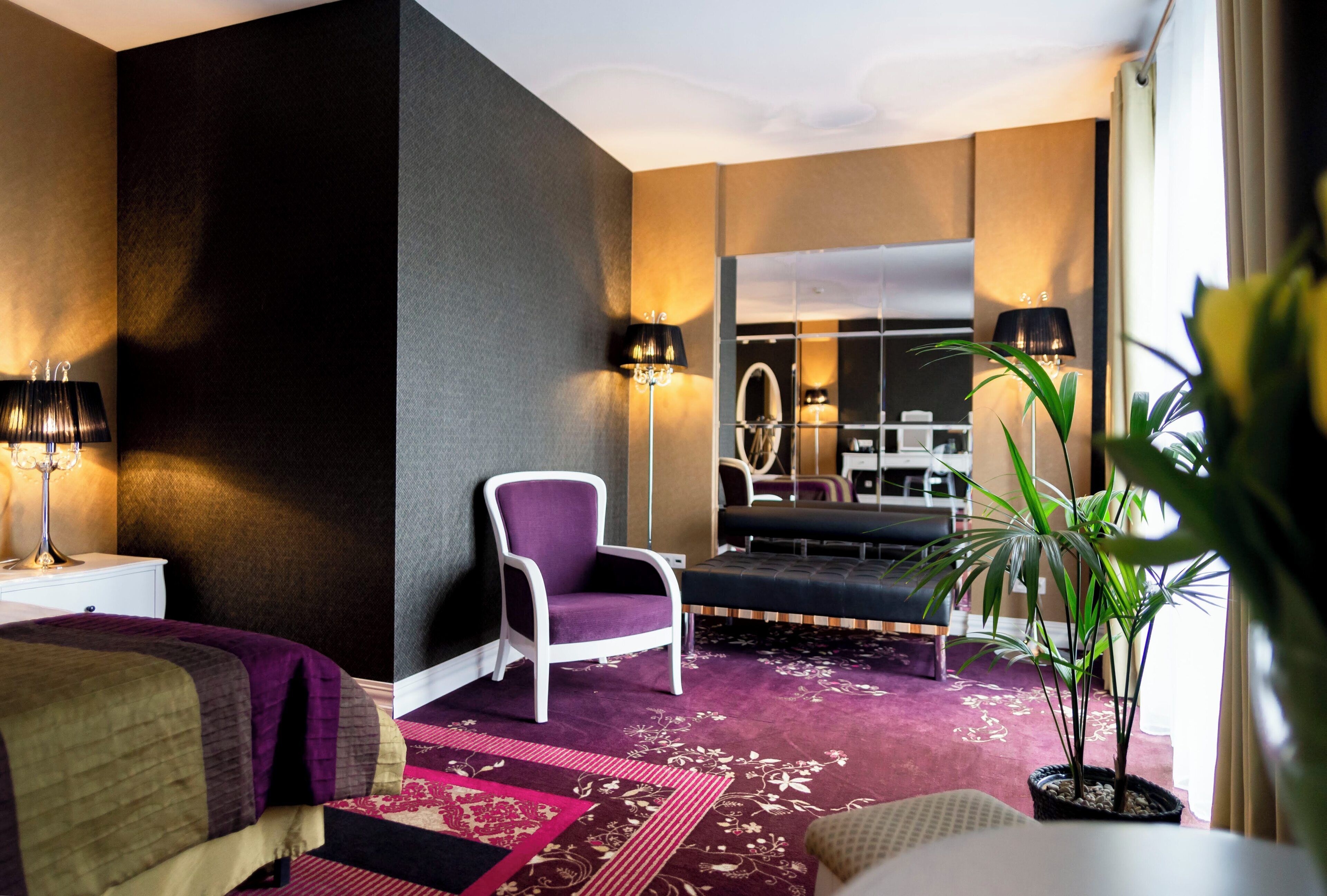 Quarto de hotel com cama, poltrona roxa, sofá, espelho grande e planta em vaso.