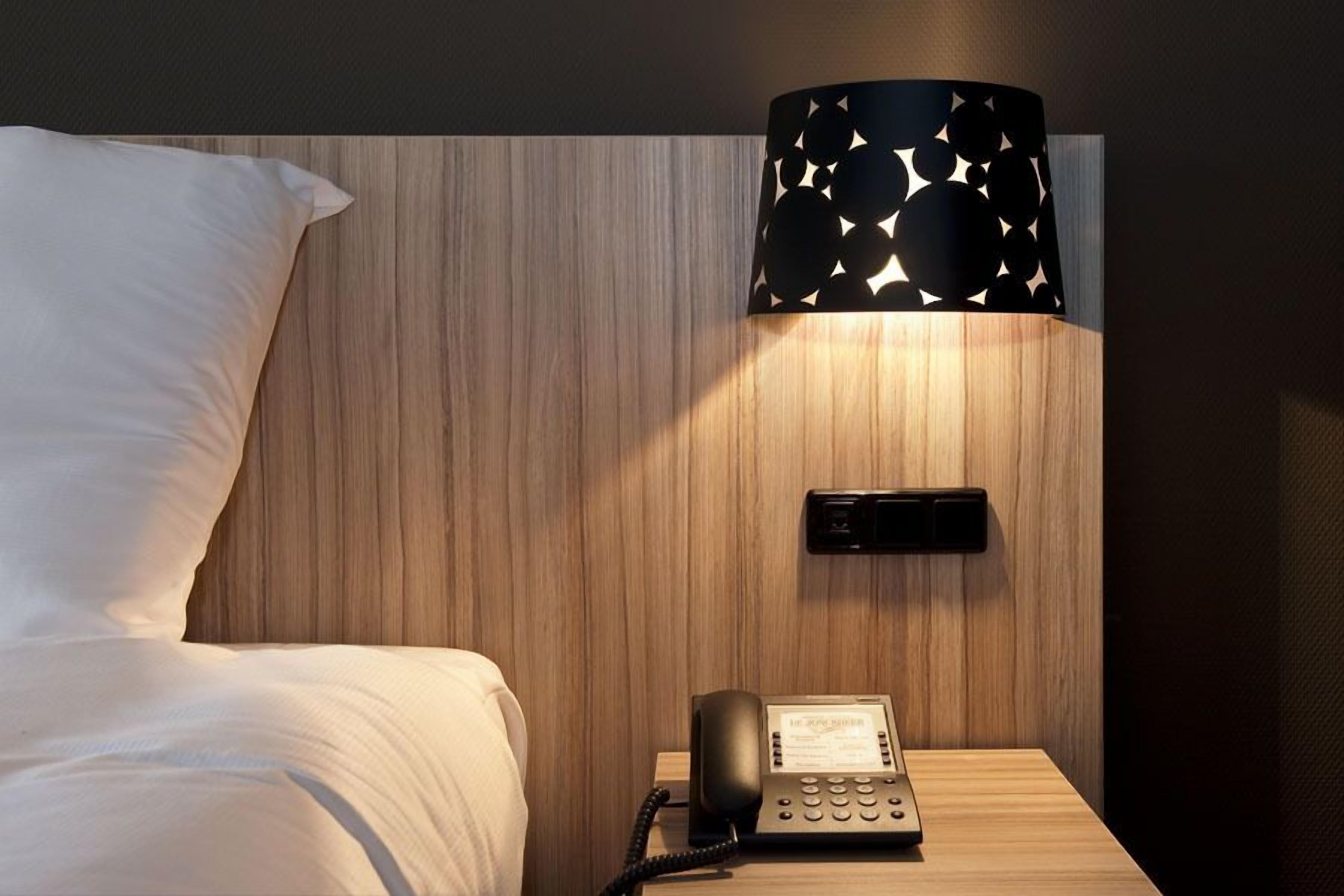 Quarto de hotel com cabeceira de madeira, travesseiro branco, mesa de cabeceira com telefone e abajur com padrão de estrela.
