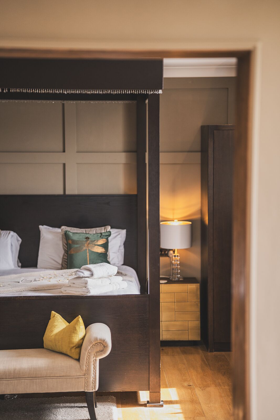 Uma cama de dossel com roupa de cama branca e um travesseiro verde, uma mesa de cabeceira com abajur e uma cadeira com almofada amarela.