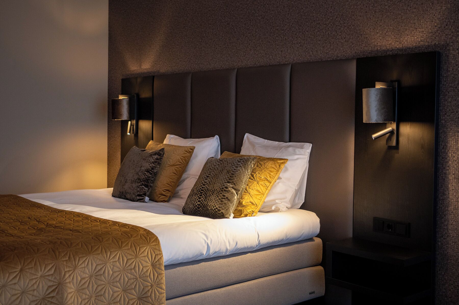 Cama bem arrumada com colcha estampada, várias almofadas e cabeceira com iluminação embutida.