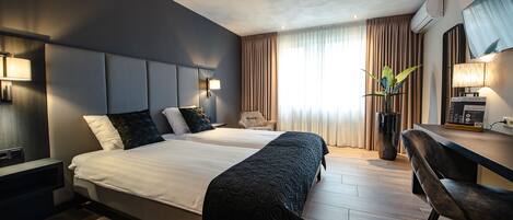 Quarto de hotel com uma cama grande, uma escrivaninha, uma cadeira, uma planta e uma televisão.