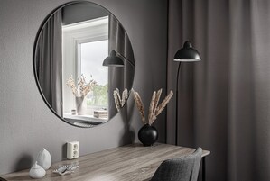 Quarto moderno com espelho redondo, escrivaninha de madeira, um vaso com plumas de capim-do-campo e duas luminárias de chão pretas.