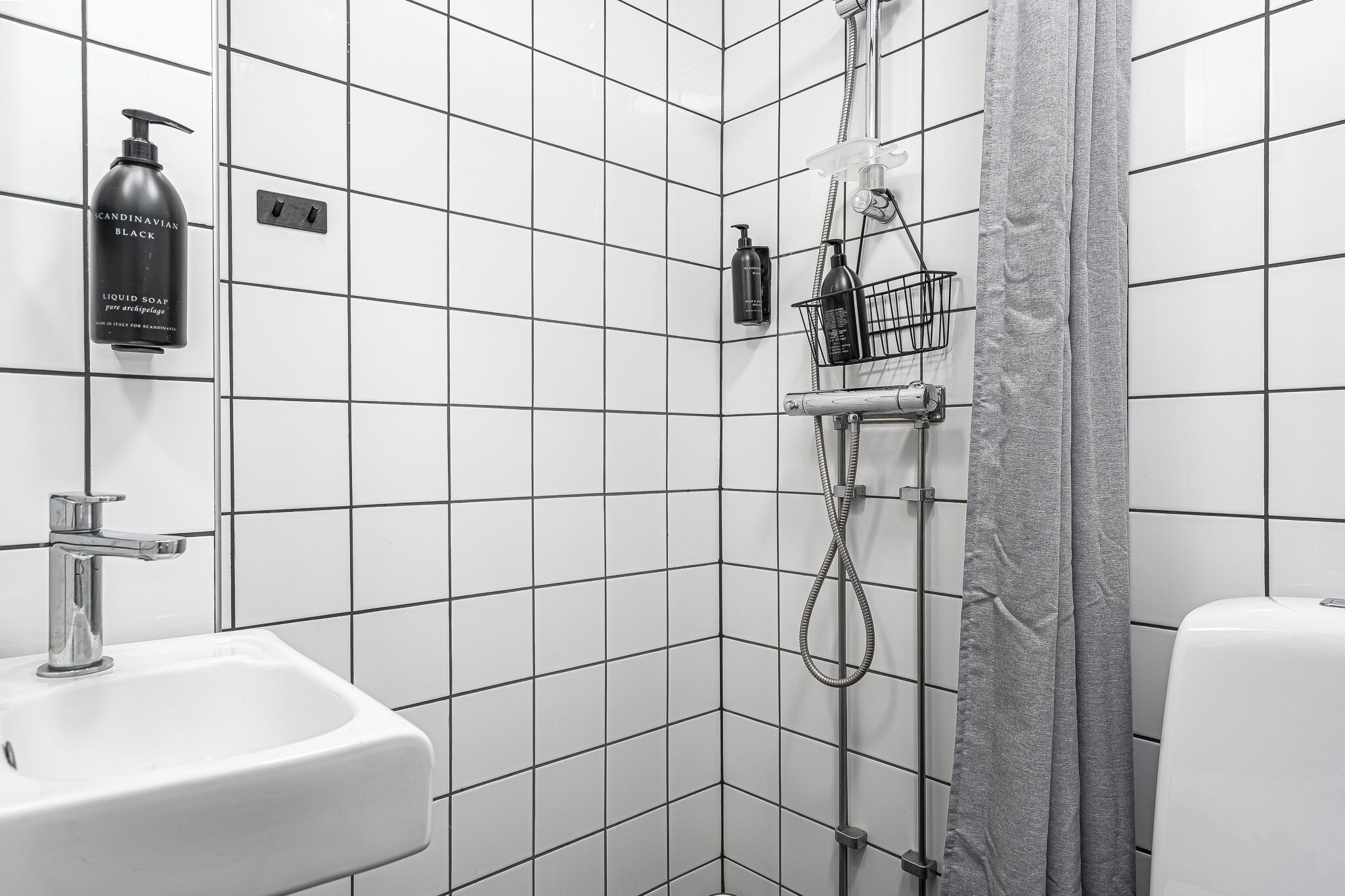 Um banheiro com paredes revestidas de azulejos brancos, pia, chuveiro com cortina cinza e artigos de higiene suspensos na parede.